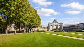 Um edifício grande e histórico com um arco e colunas, cercado por jardins bem cuidados e árvores.