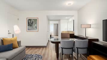 Quarto de hotel moderno com sofá, mesa de jantar, duas cadeiras, TV e banheiro visível através de uma porta aberta.