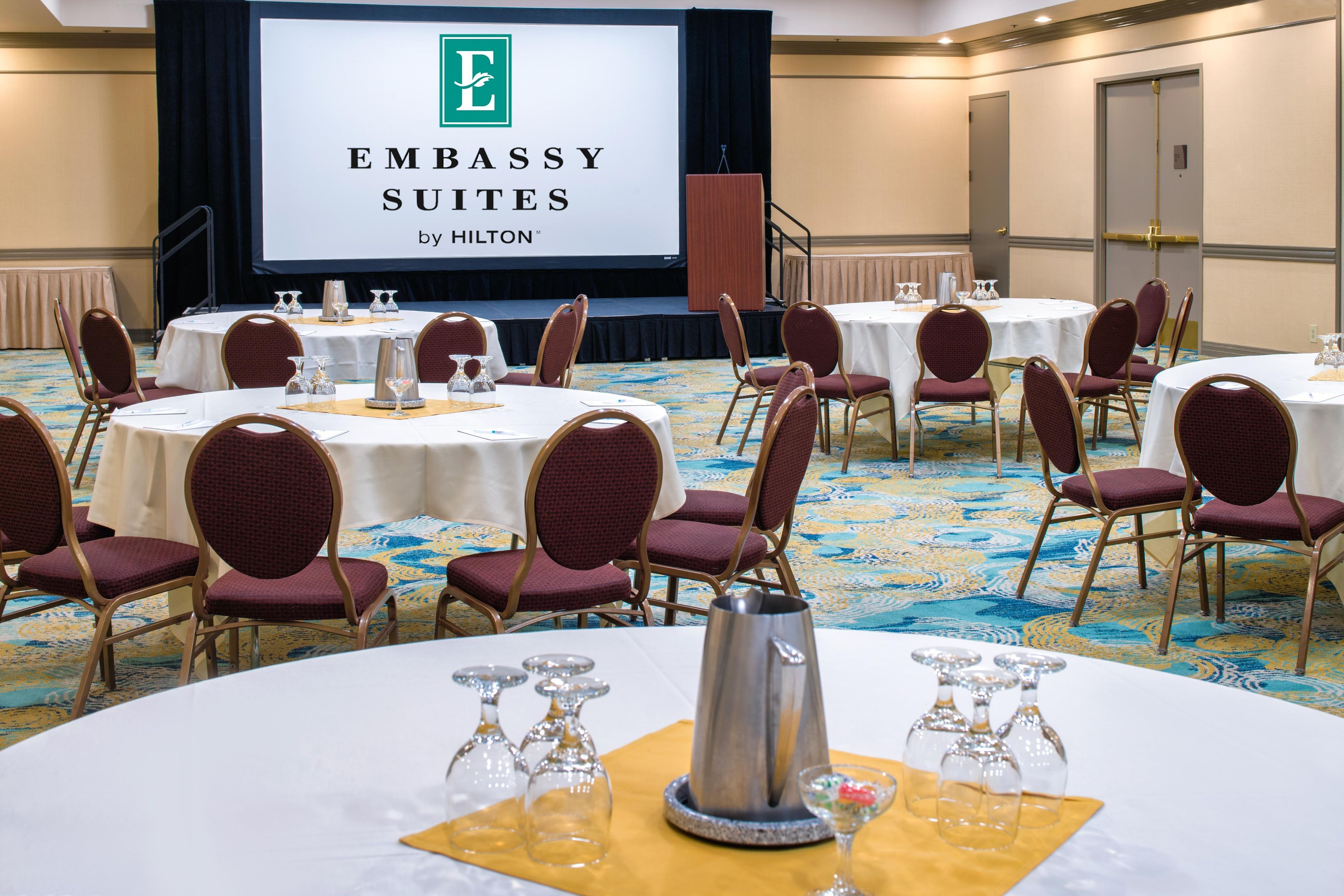 Uma sala de conferências com mesas redondas, cadeiras e uma tela exibindo 'EMBASSY SUITES by HILTON'.
