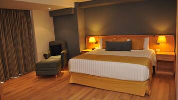 Quarto de hotel com uma cama grande, duas mesas de cabeceira com luminárias, uma cadeira e piso de madeira.