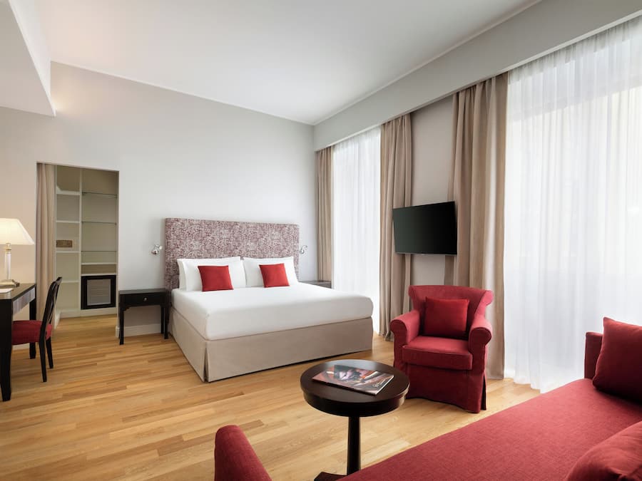 Quarto de hotel com cama, poltrona vermelha, sofá, televisão, escrivaninha com cadeira e mesa pequena.