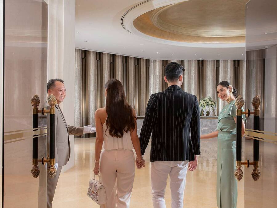 Um grupo de pessoas caminhando por um hall moderno de hotel, com piso polido, teto curvo e um espelho grande refletindo o interior.
