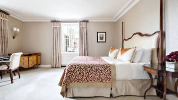 Quarto de hotel com uma cama grande, uma escrivaninha e uma cadeira. Há uma janela com cortinas, um quadro na parede e um vaso com flores sobre uma mesinha de cabeceira.