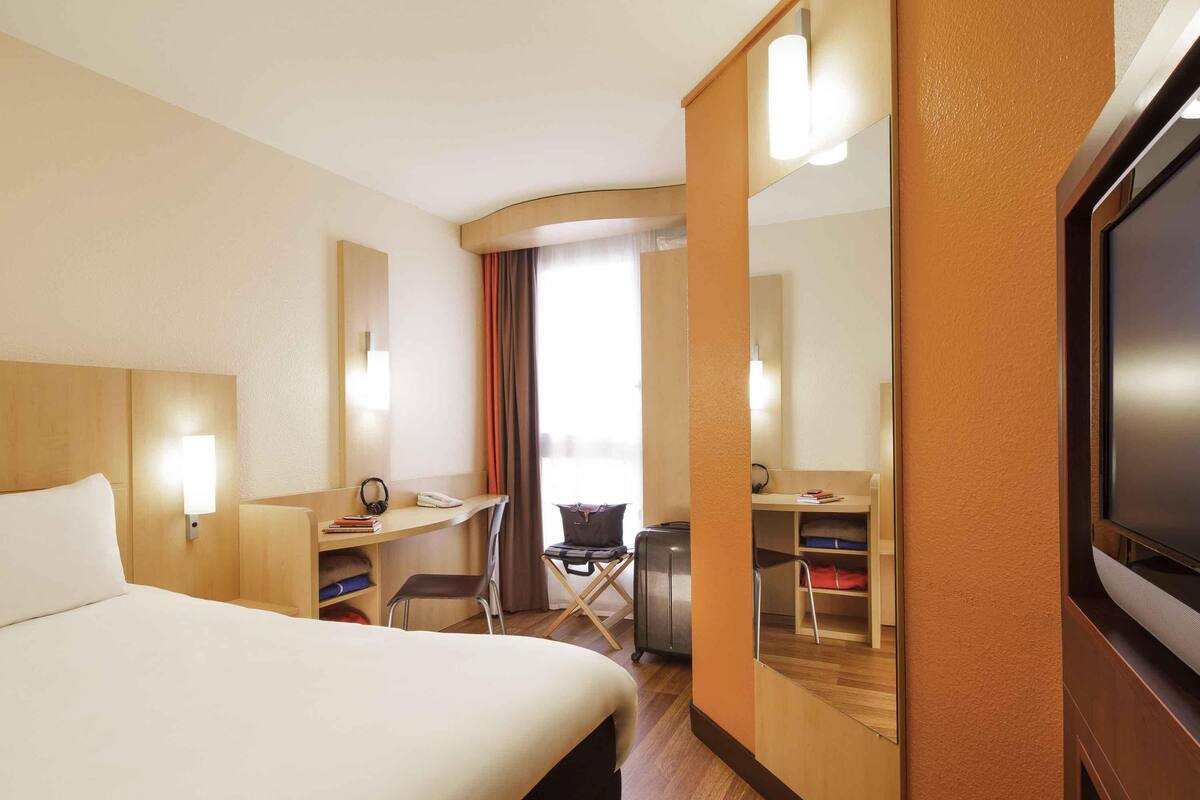 Quarto de hotel com cama, escrivaninha, cadeira e televisão.