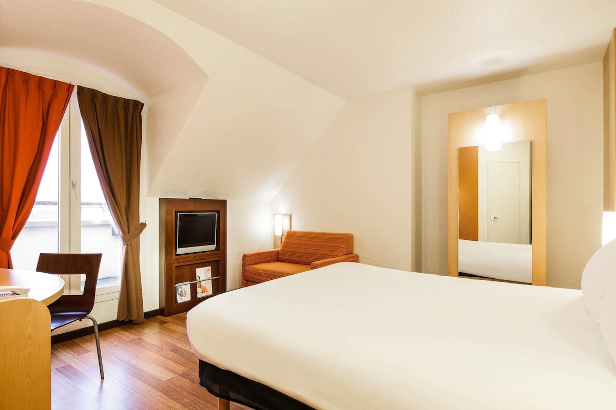Quarto de hotel com cama, escrivaninha, cadeira, televisão e espelho.
