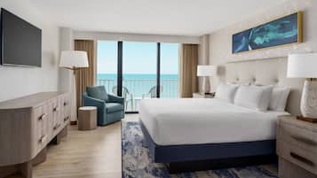 Quarto de hotel com uma cama grande, uma escrivaninha, uma cadeira, uma mesinha de apoio e varanda com vista para o mar.