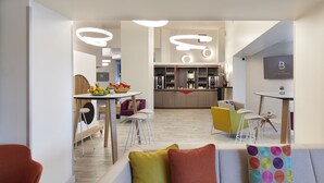 Um interior moderno com um sofá, almofadas coloridas, uma área de refeições com mesa e cadeiras, e uma cozinha com geladeira e bancada.