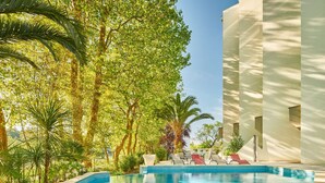 Uma área de piscina com piscina azul cristalina, rodeada por vegetação exuberante e palmeiras. Há espreguiçadeiras e uma mesa perto da piscina.