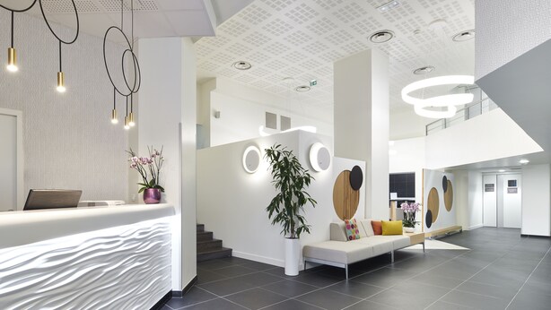 Uma área de recepção moderna com um balcão curvo, um espaço de estar com sofá e almofadas, e uma grande planta em vaso.