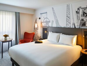 Quarto de hotel com uma cama grande, uma poltrona vermelha, uma mesa pequena com um vaso de flores e um mural na parede com a figura de garças.