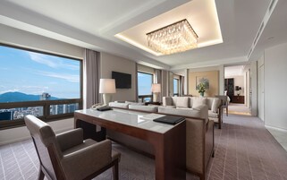 Quarto de hotel com uma grande janela, um candelabro, um conjunto de sofás, uma escrivaninha e uma televisão de tela plana.