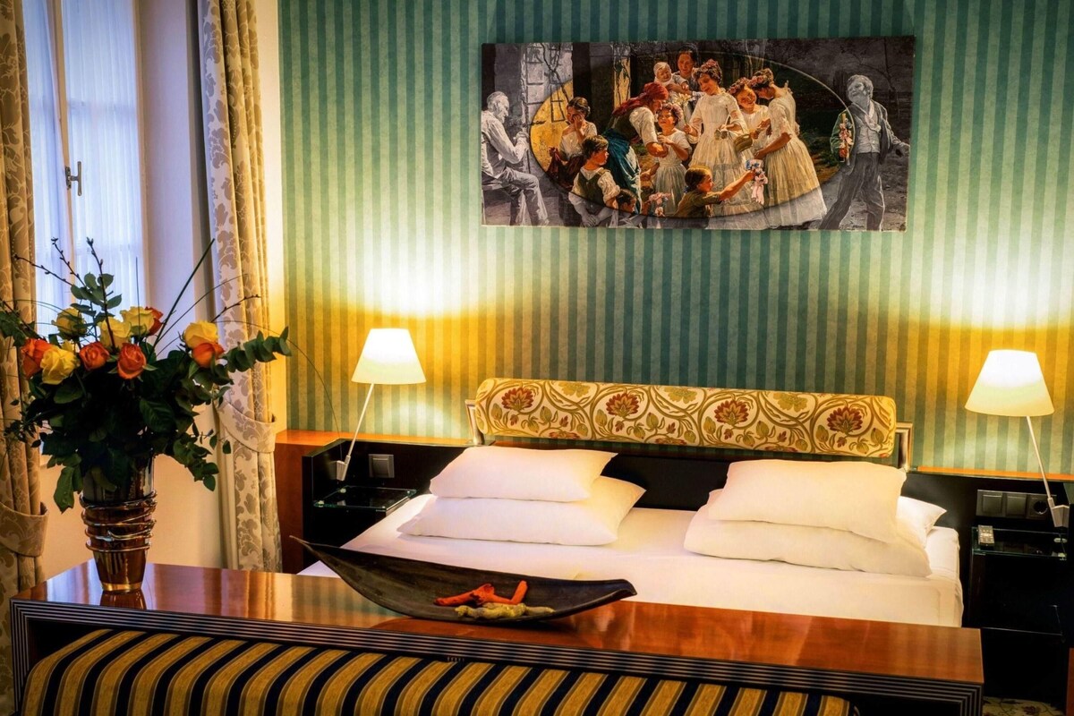Quarto de hotel com cama, duas luminárias de cabeceira, um vaso com flores e um quadro na parede.