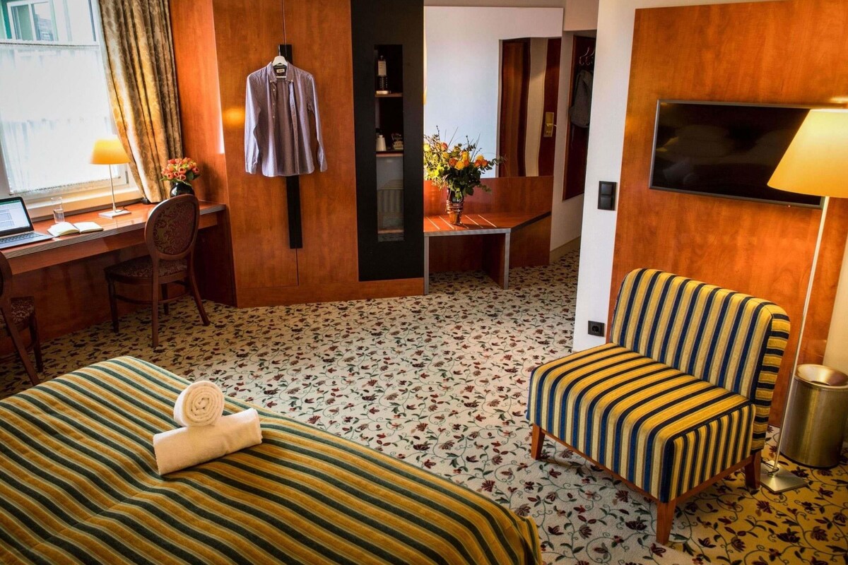 Quarto de hotel com cama, secretária, cadeira, televisão e pequena mesa com um vaso de flores.
