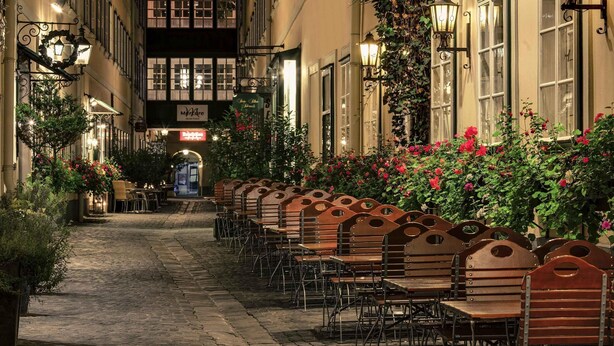 Uma rua de paralelepípedos com bancos e mesas de madeira, um café com letreiro e edifícios com grandes janelas.