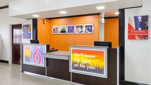 Balcão de recepç ão de hotel com a placa 'Motel 6', uma placa com a frase 'Deixamos a luz acesa para você' e um cartaz de 'Reserve Direto e Economize'.