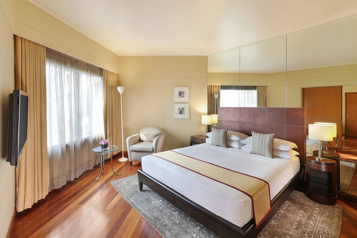 Quarto de hotel com uma cama grande, uma cadeira, uma mesa pequena, uma TV e um espelho.