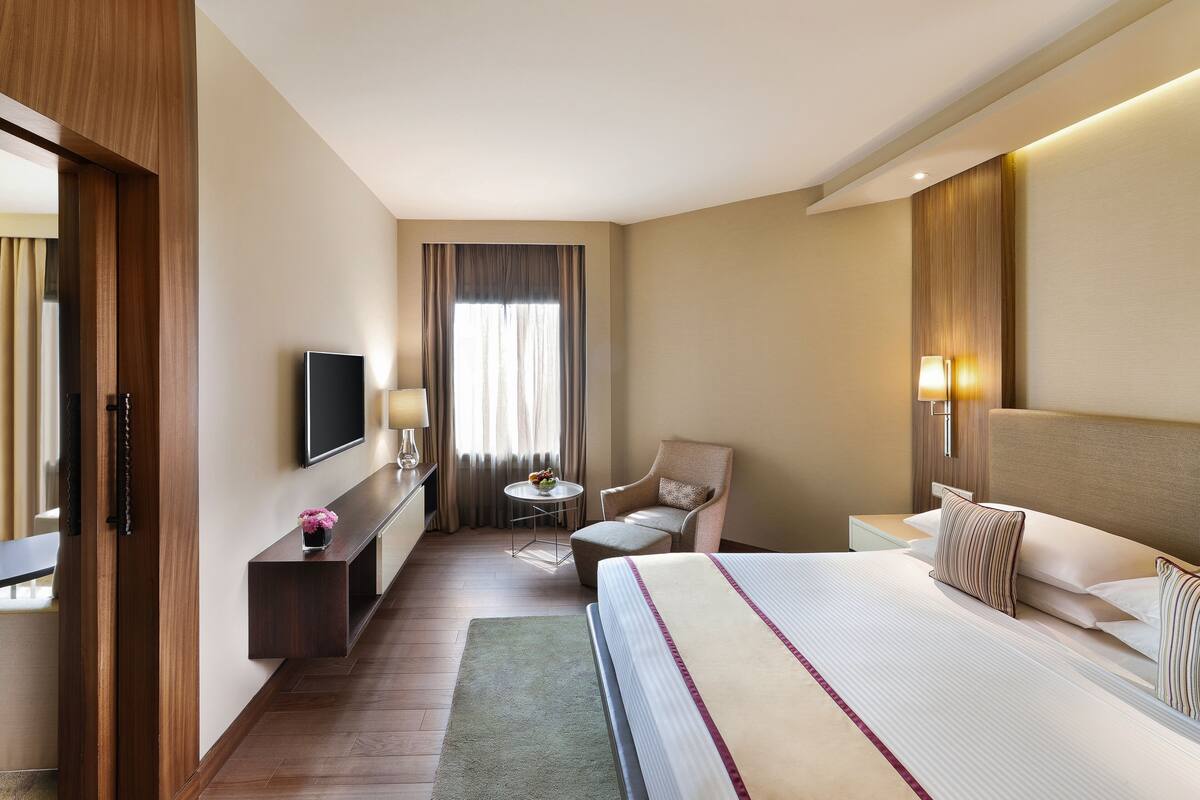 Quarto de hotel com uma cama grande, televisão, secretária com luminária, cadeira e pequena mesa com um vaso de flores.