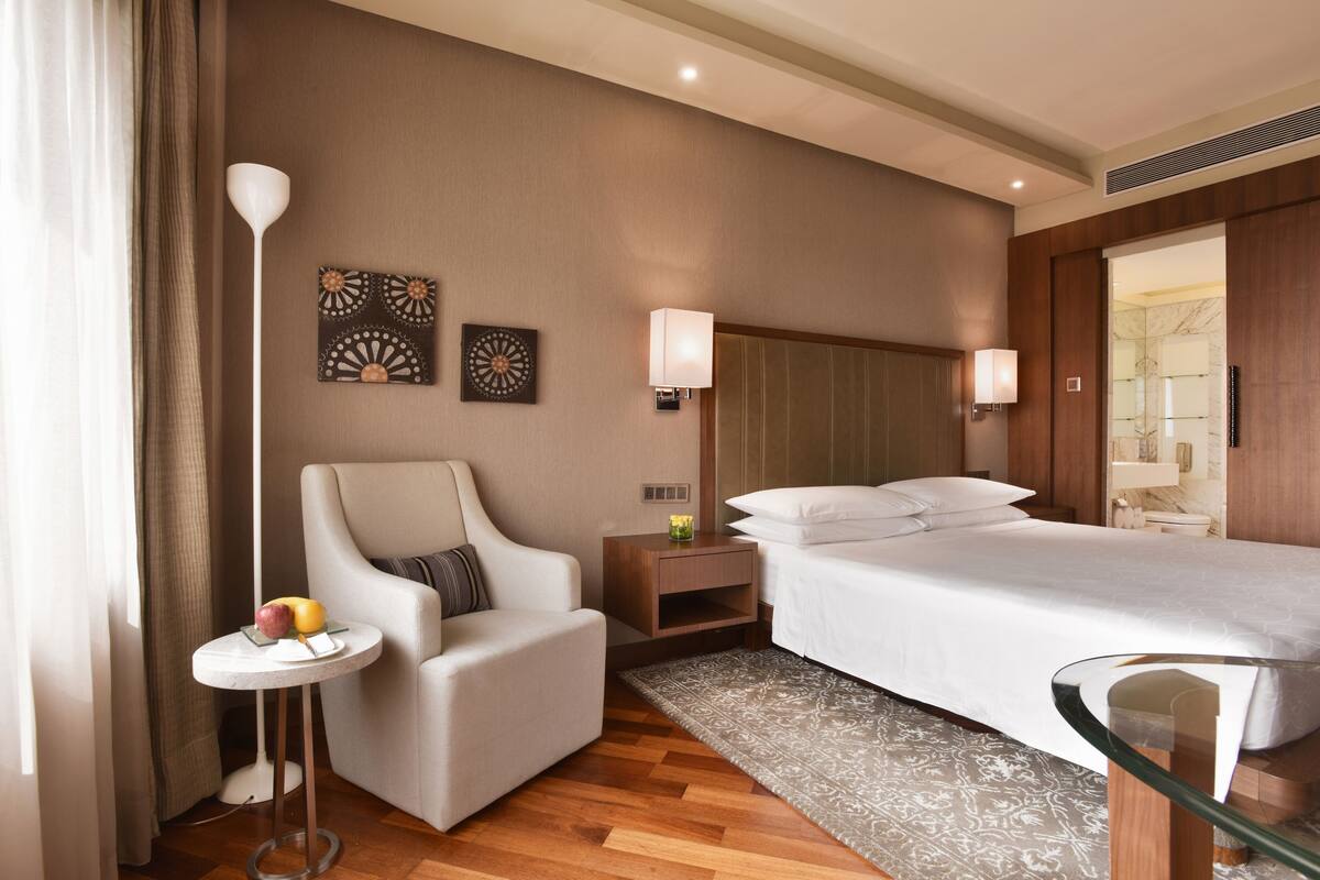 Quarto de hotel com cama, uma cadeira, uma mesinha com frutas, abajur e decoração na parede.