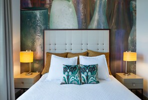 Uma cama bem arrumada com lençóis brancos, dois travesseiros decorativos e duas luminárias de cabeceira.