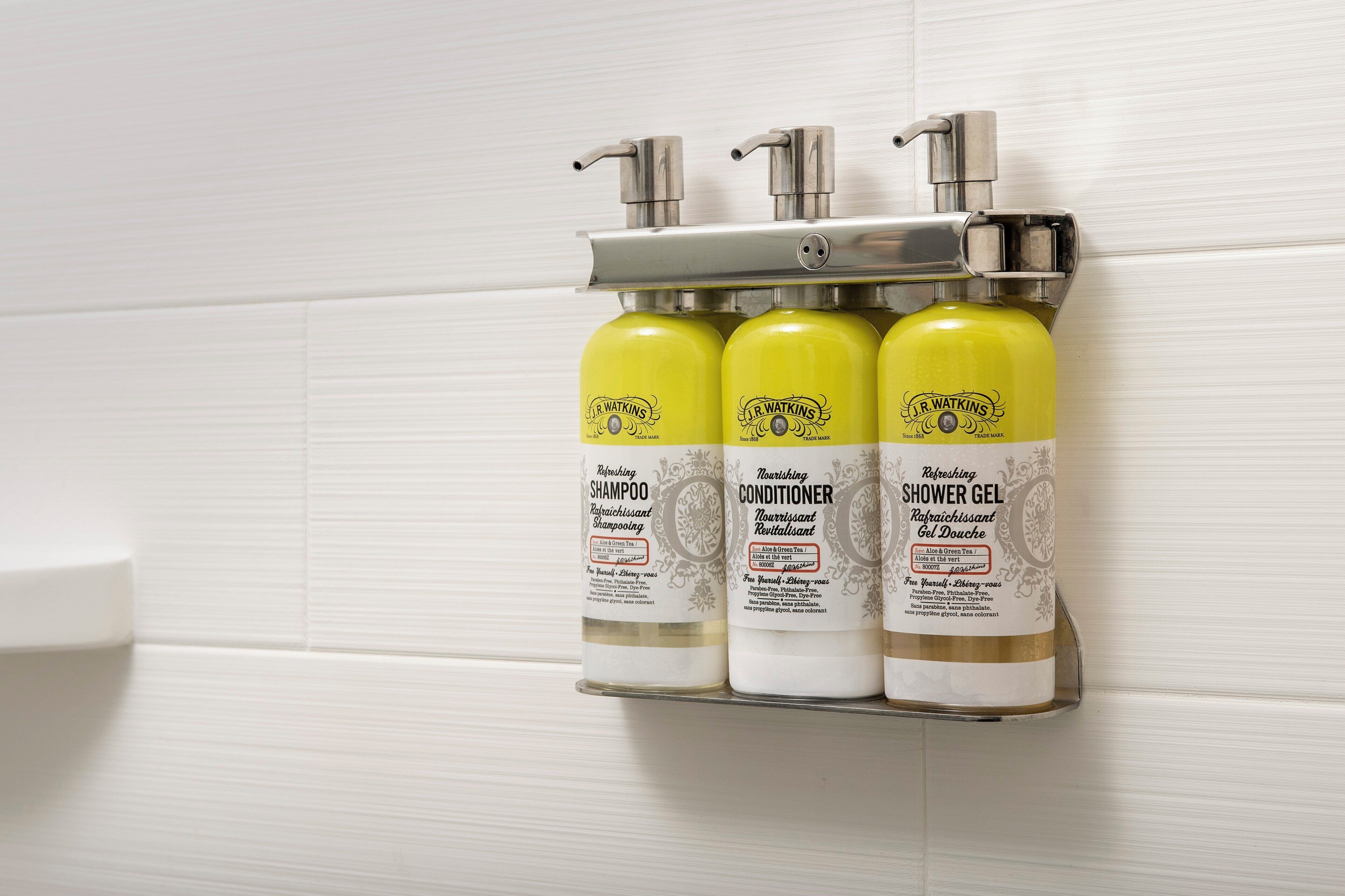 Casa de banho com três garrafas amarelas de shampoo, condicionador e gel de banho J.R. Watkins fixadas numa parede.