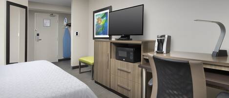 Quarto de hotel com cama, escrivaninha com computador, micro-ondas, televisão de tela plana e uma cadeira.