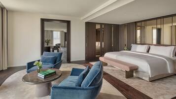 Quarto de hotel moderno com uma cama grande, área de estar com sofá e poltronas, mesa de centro e um espelho amplo.