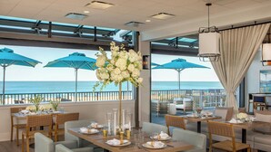 Um restaurante com mesas preparadas para refeições, grandes janelas com vista para o oceano e guarda-sóis azuis.