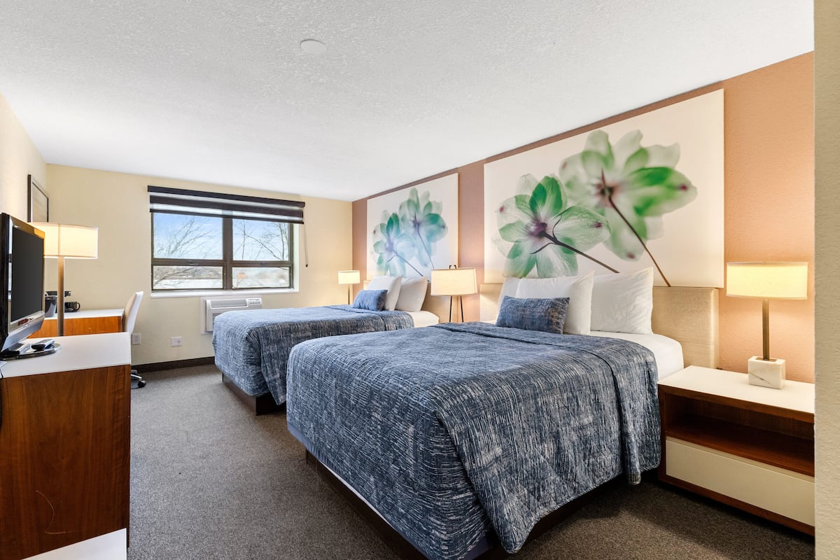 Quarto de hotel com duas camas, uma mesa com computador, televisão e obras de arte nas paredes.