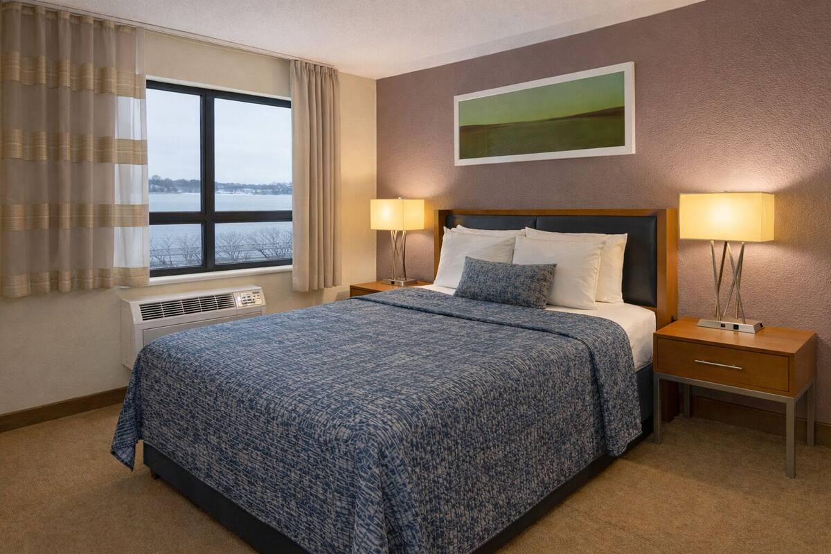 Quarto de hotel com uma cama grande, duas luminárias de cabeceira, uma janela com cortinas, um quadro na parede e um aparelho de ar condicionado.