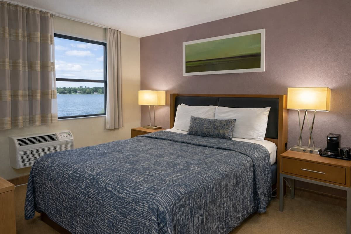 Quarto de hotel com cama, duas mesas de cabeceira, janela com cortinas, ar condicionado, um quadro na parede e vista para a água.