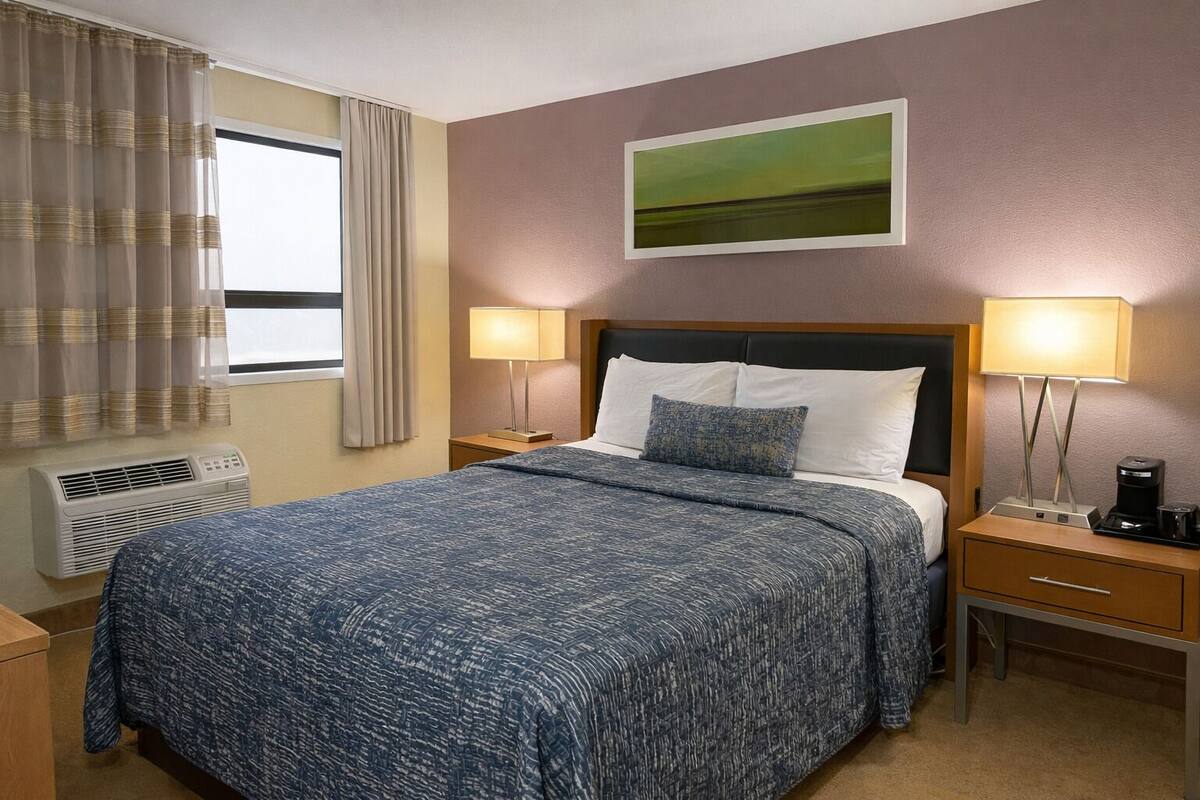 Quarto de hotel com cama, duas mesas de cabeceira, luminária na parede, janela com cortinas e aparelho de ar condicionado.