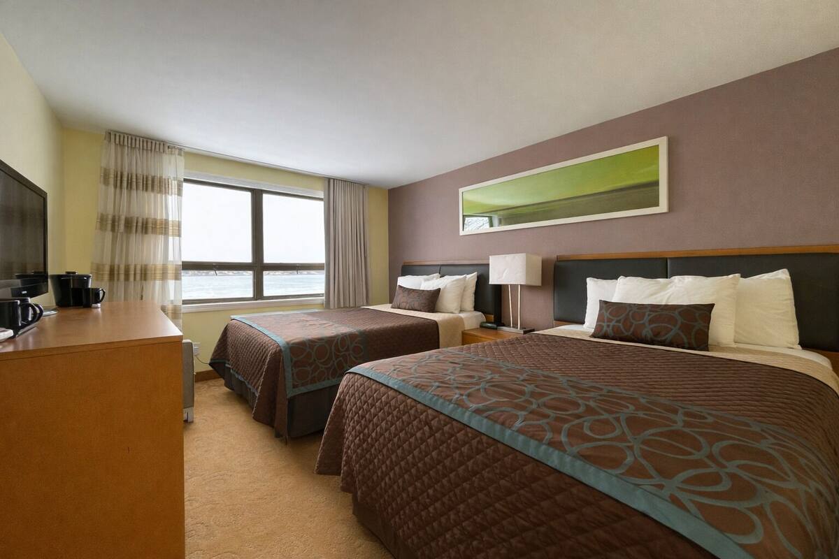 Quarto de hotel com duas camas, uma escrivaninha, televisão e janela com cortinas.