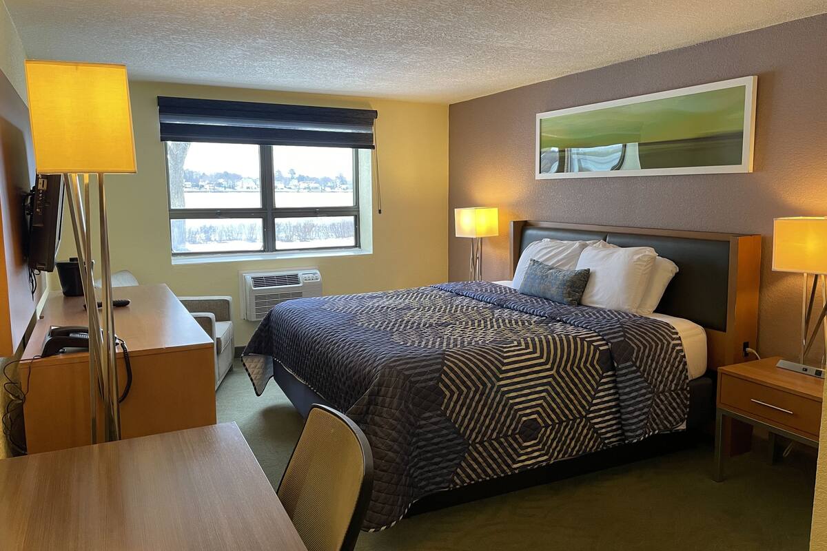 Quarto de hotel com cama, abajures, escrivaninha com telefone, cadeira, janela com vista e um quadro na parede.
