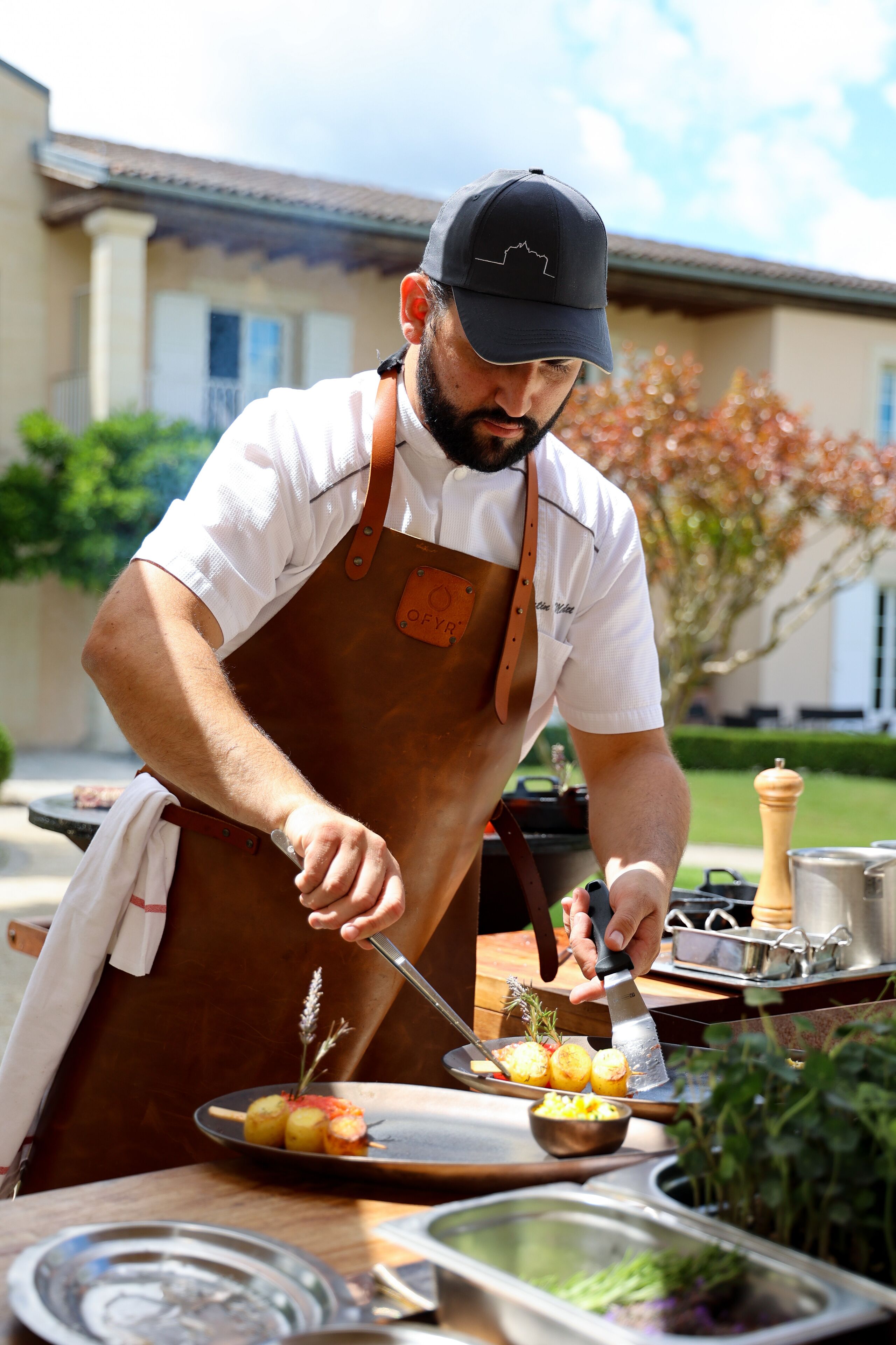Um chef preparando comida ao ar livre, com uma churrasqueira e diversos utensílios de cozinha.