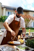 Um chef preparando comida ao ar livre, com uma churrasqueira e diversos utensílios de cozinha.