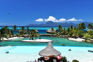 Resort com piscina, palmeiras e vista para o oceano e montanhas.