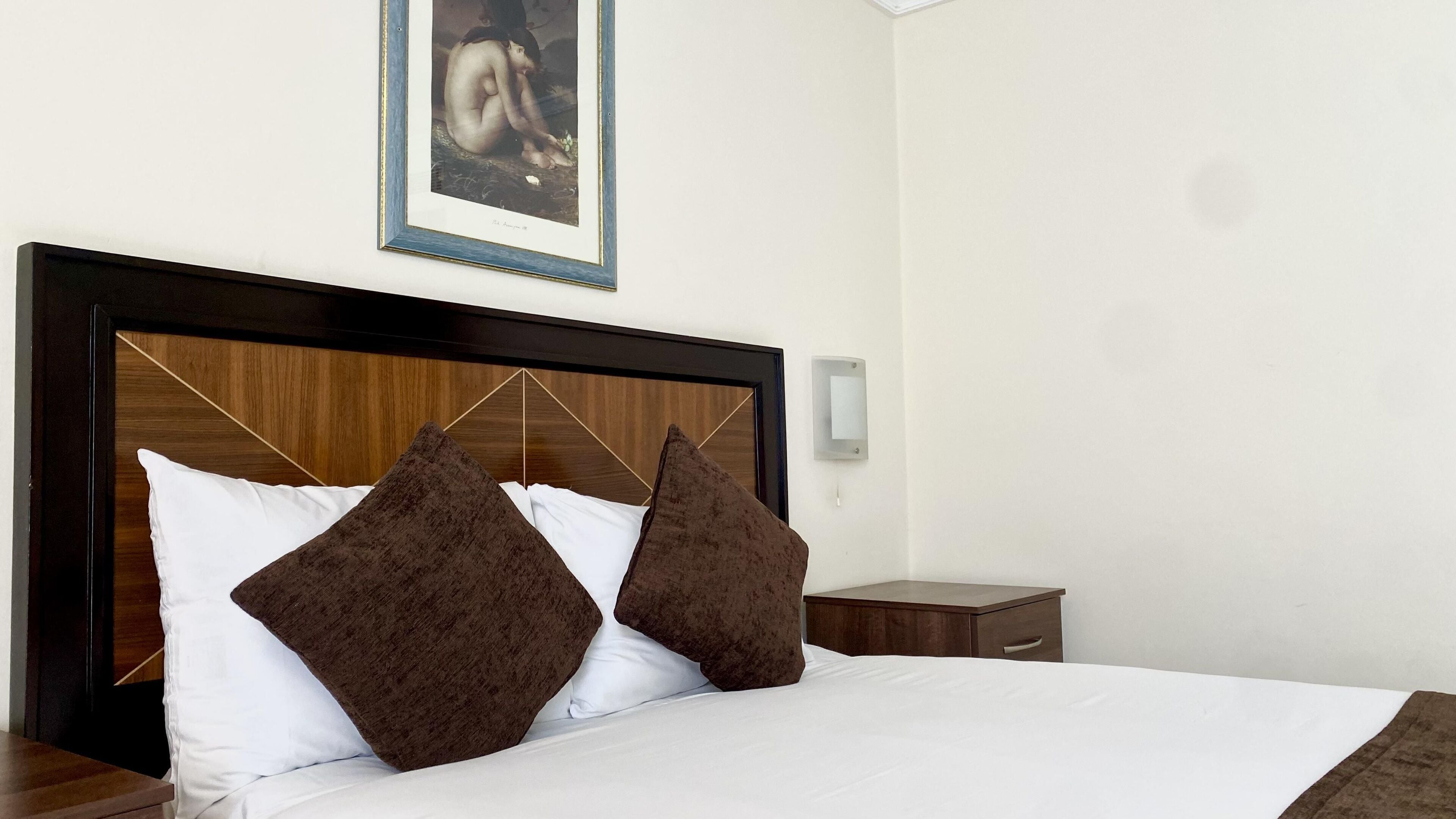Uma cama bem arrumada com almofadas brancas e castanhas, uma cabeceira de madeira e uma pequena mesa de cabeceira com uma luminária.