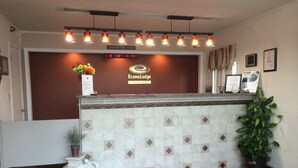 Uma área de recepção de hotel, com balcão de check-in, painel digital e tapete vermelho com o nome do hotel.