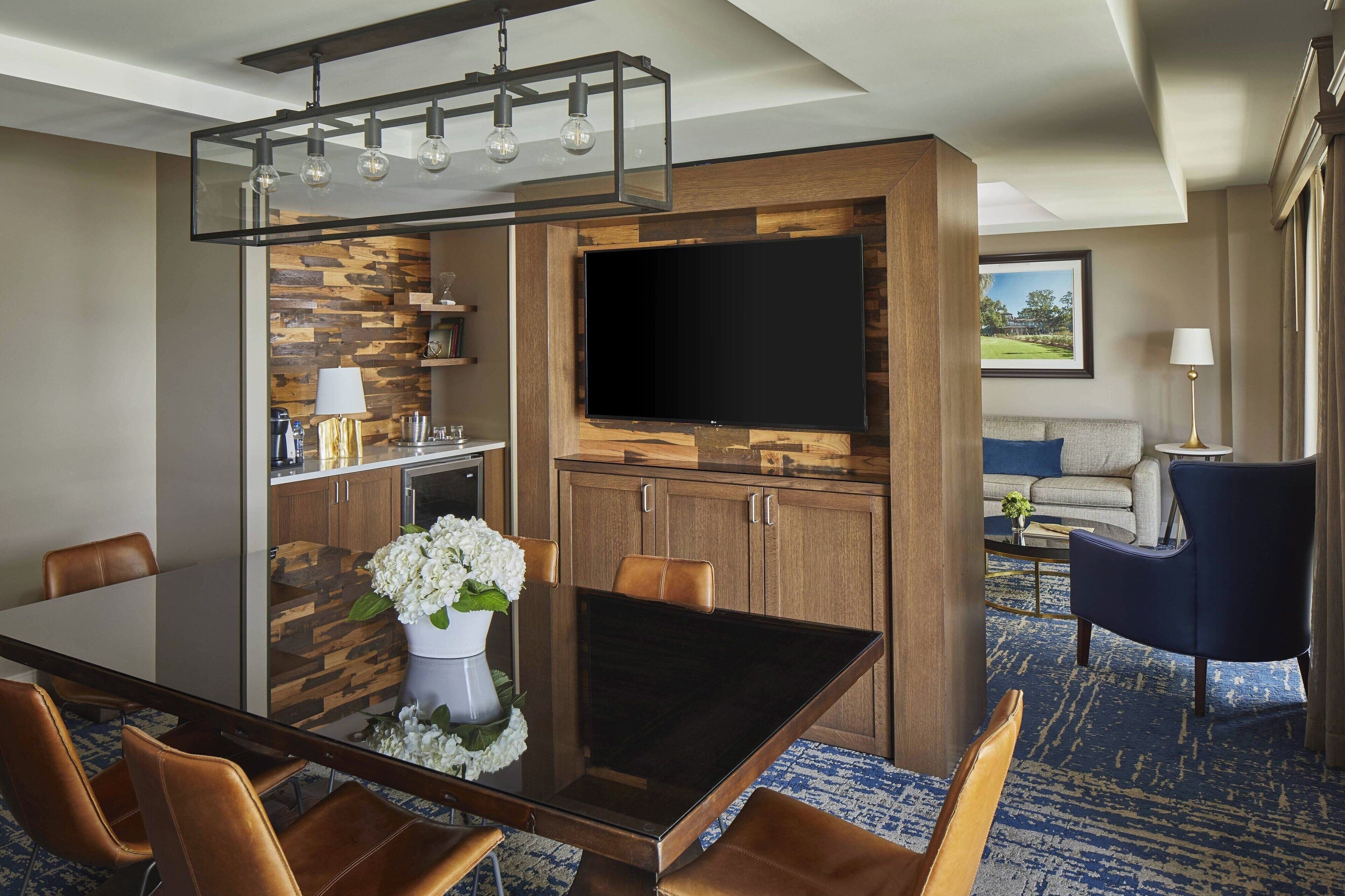 Quarto de hotel moderno com área de refeições, cozinha, TV de tela plana e canto de estar com uma poltrona azul.