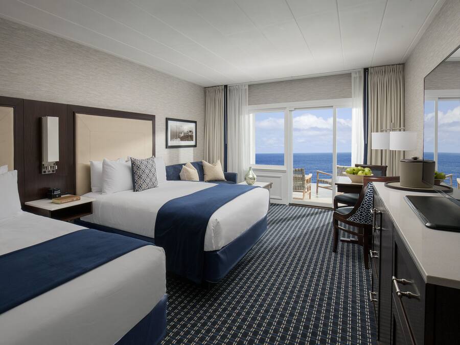 Quarto de hotel com duas camas, uma escrivaninha com luminária, uma cadeira e vista para o oceano.