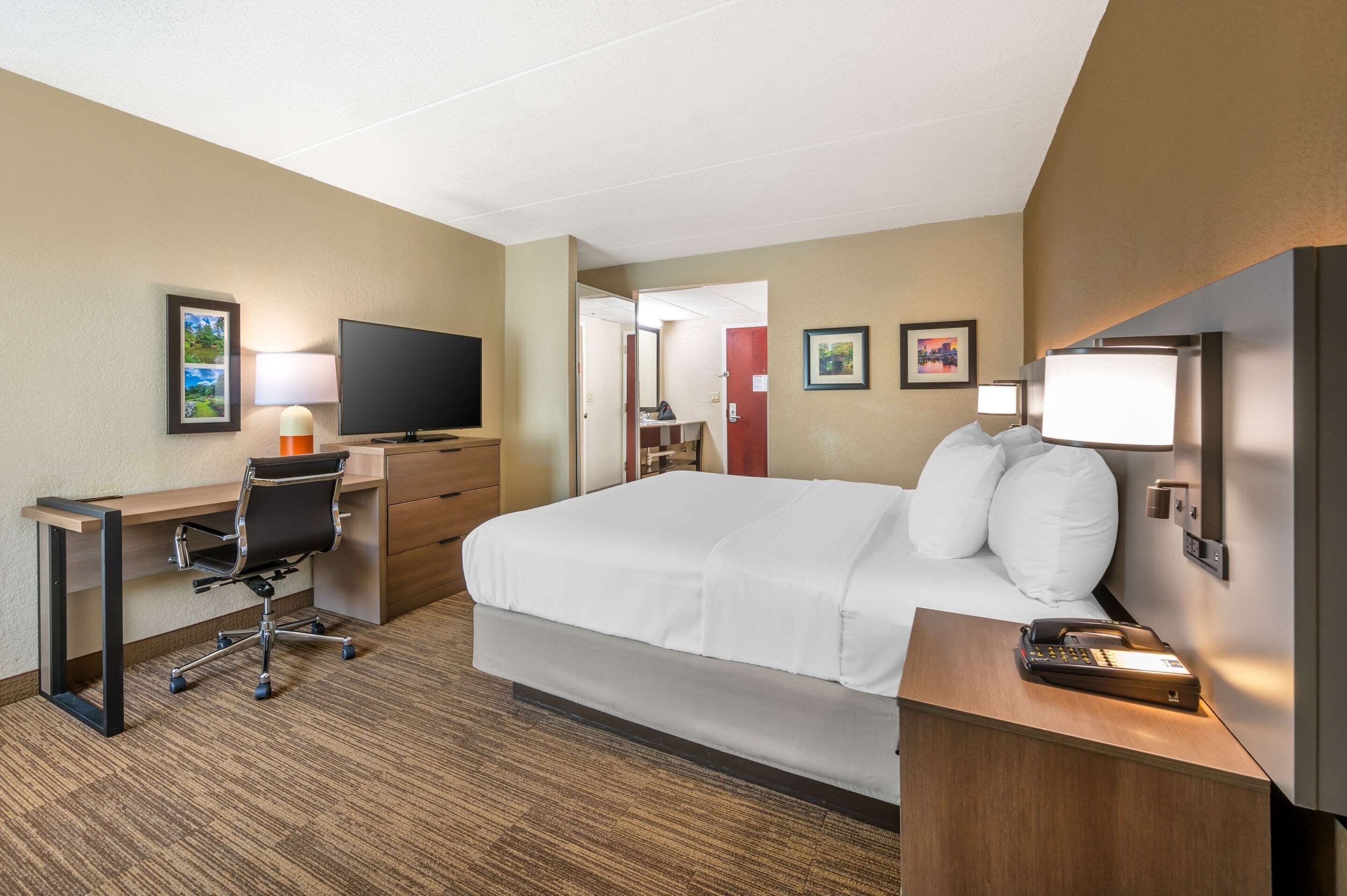 Quarto de hotel com cama, escrivaninha, cadeira, televisão e mesa de cabeceira com abajur.