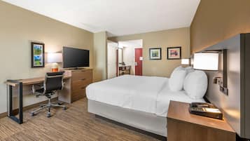 Quarto de hotel com cama, escrivaninha, cadeira, televisão e mesa de cabeceira com abajur.