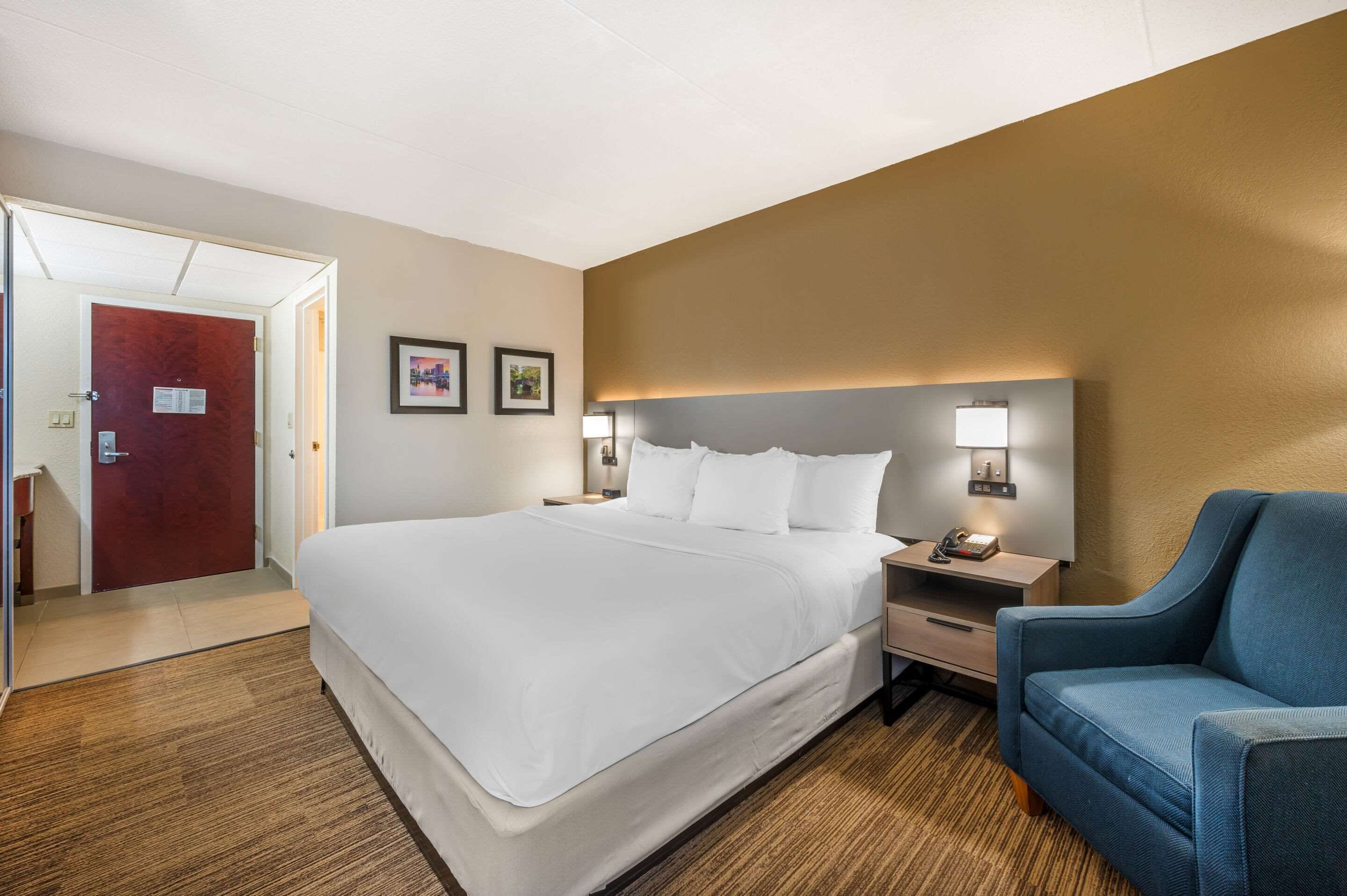Quarto de hotel com uma cama grande, uma escrivaninha com abajur, uma cadeira e duas quadros nas paredes.