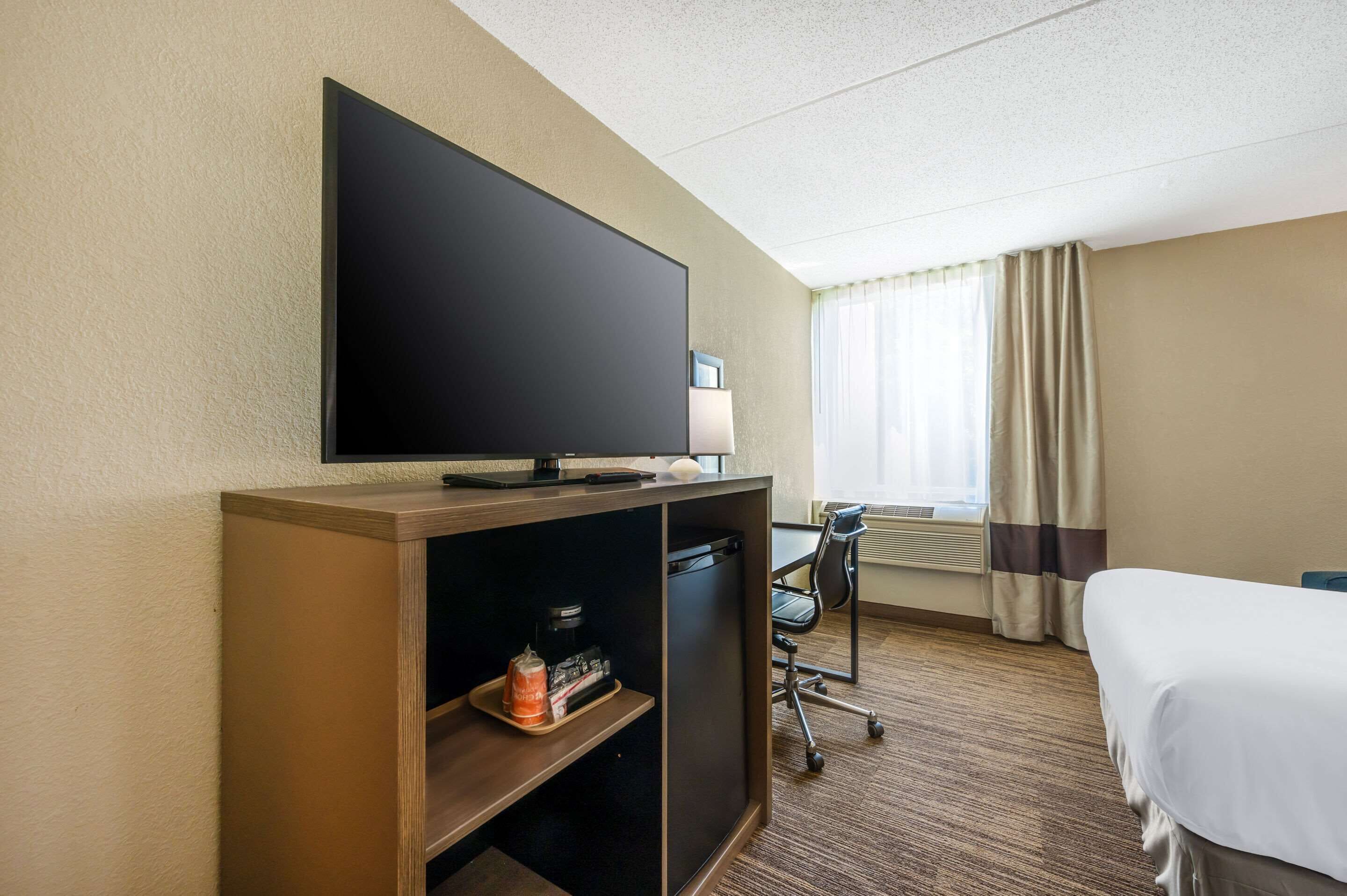 Quarto de hotel com televisão de tela plana em um suporte de madeira, uma mesa com cadeira, cama e janela com cortinas.