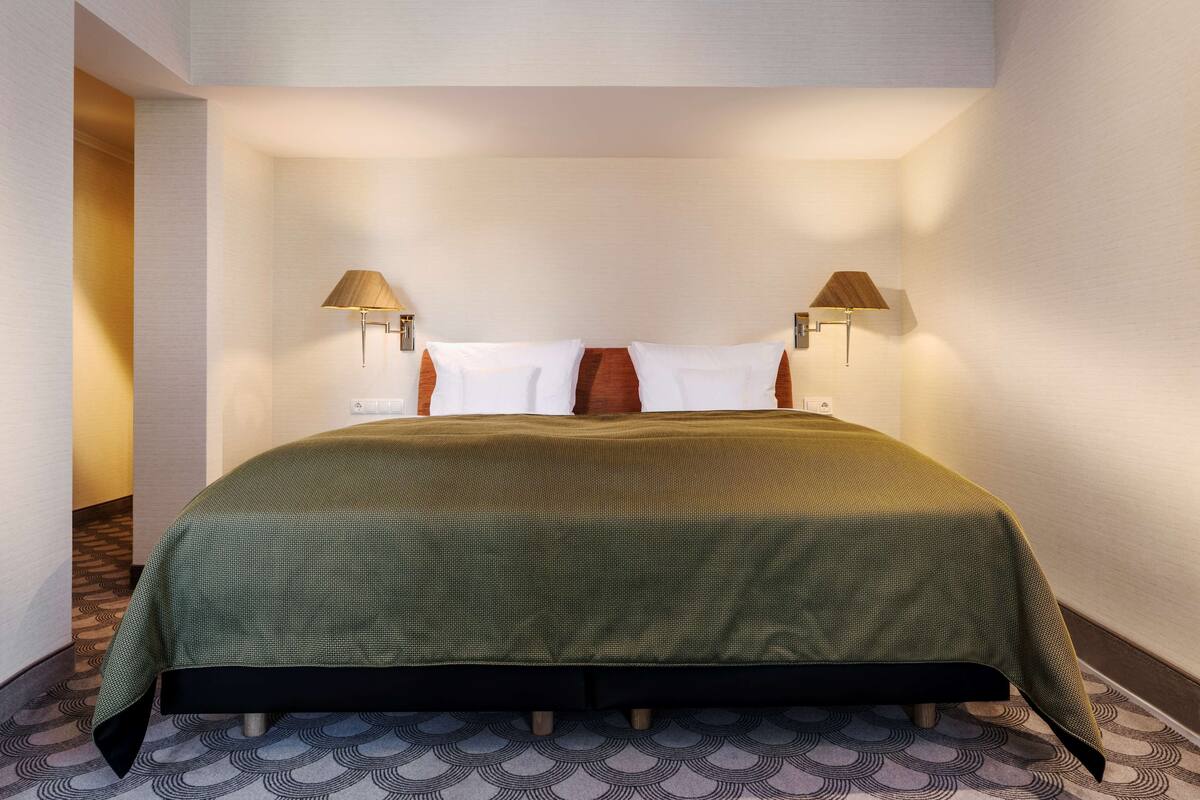 Cama bem arrumada com colcha verde, duas luminárias fixadas na parede e um tapete estampado.
