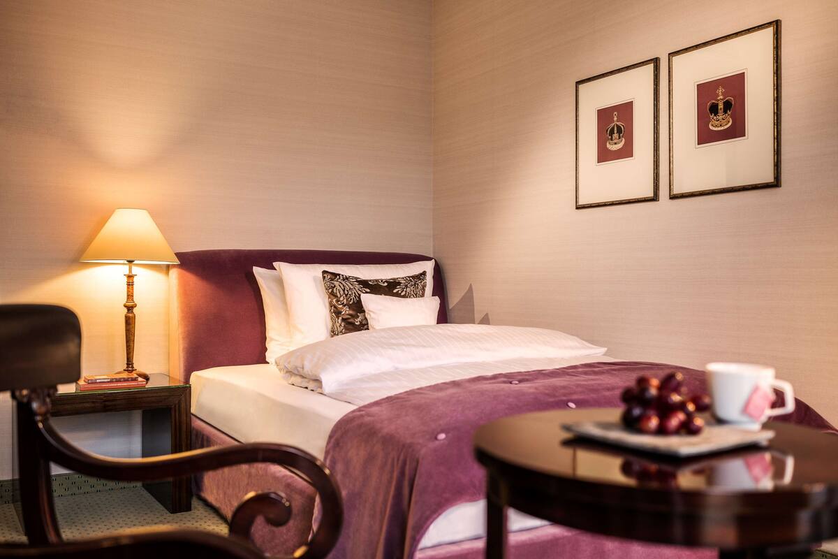 Quarto de hotel com cama, mesa de cabeceira com abajur, cadeira, pequena mesa com uma chávena e uvas, e duas molduras na parede.
