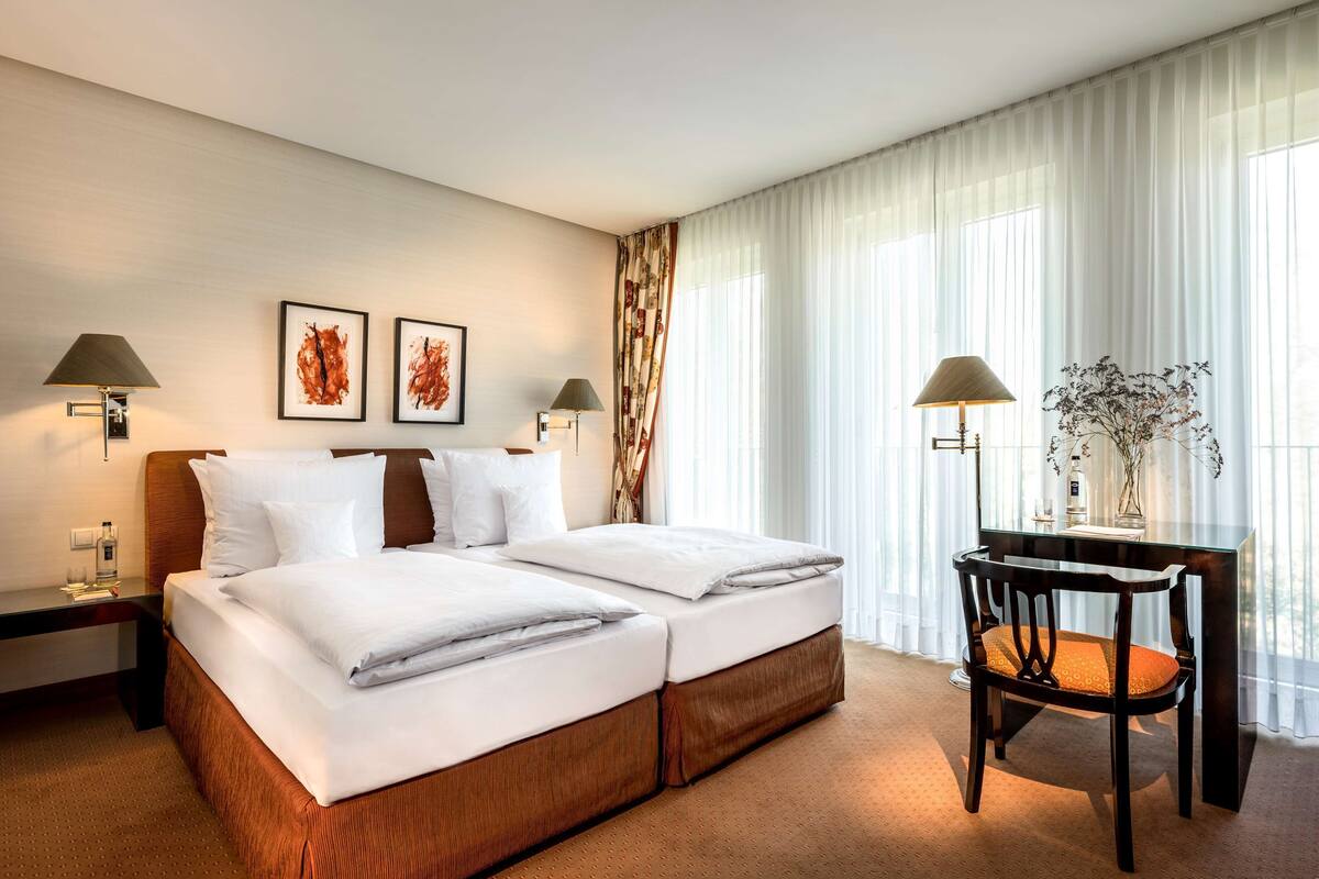 Quarto de hotel com cama de casal, duas mesas de cabeceira com luminárias, uma escrivaninha com cadeira e uma janela grande com cortinas translúcidas.