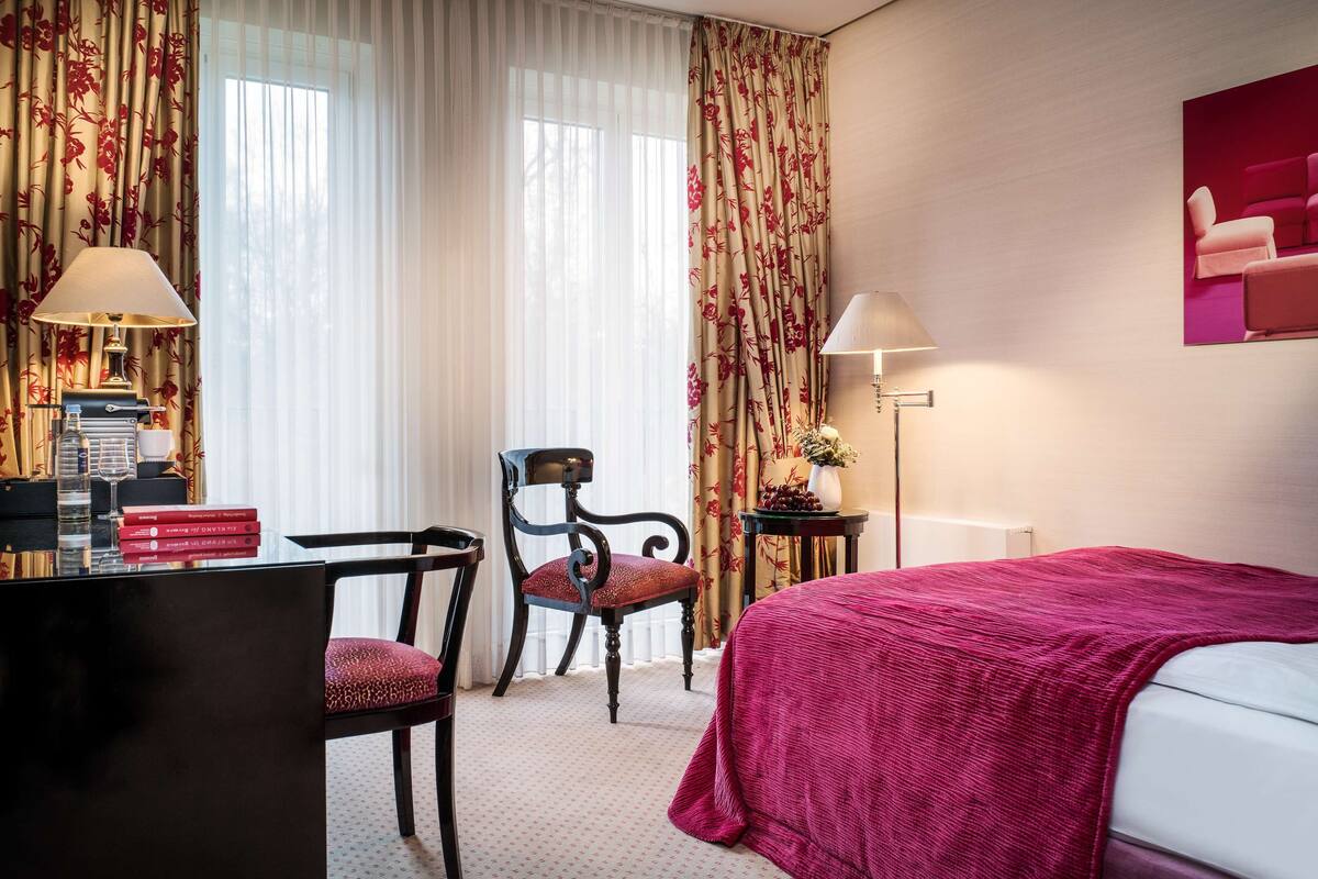 Quarto de hotel com cama, escrivaninha com luminária, cadeira, mesa pequena com um vaso e cortinas estampadas com motivos florais.