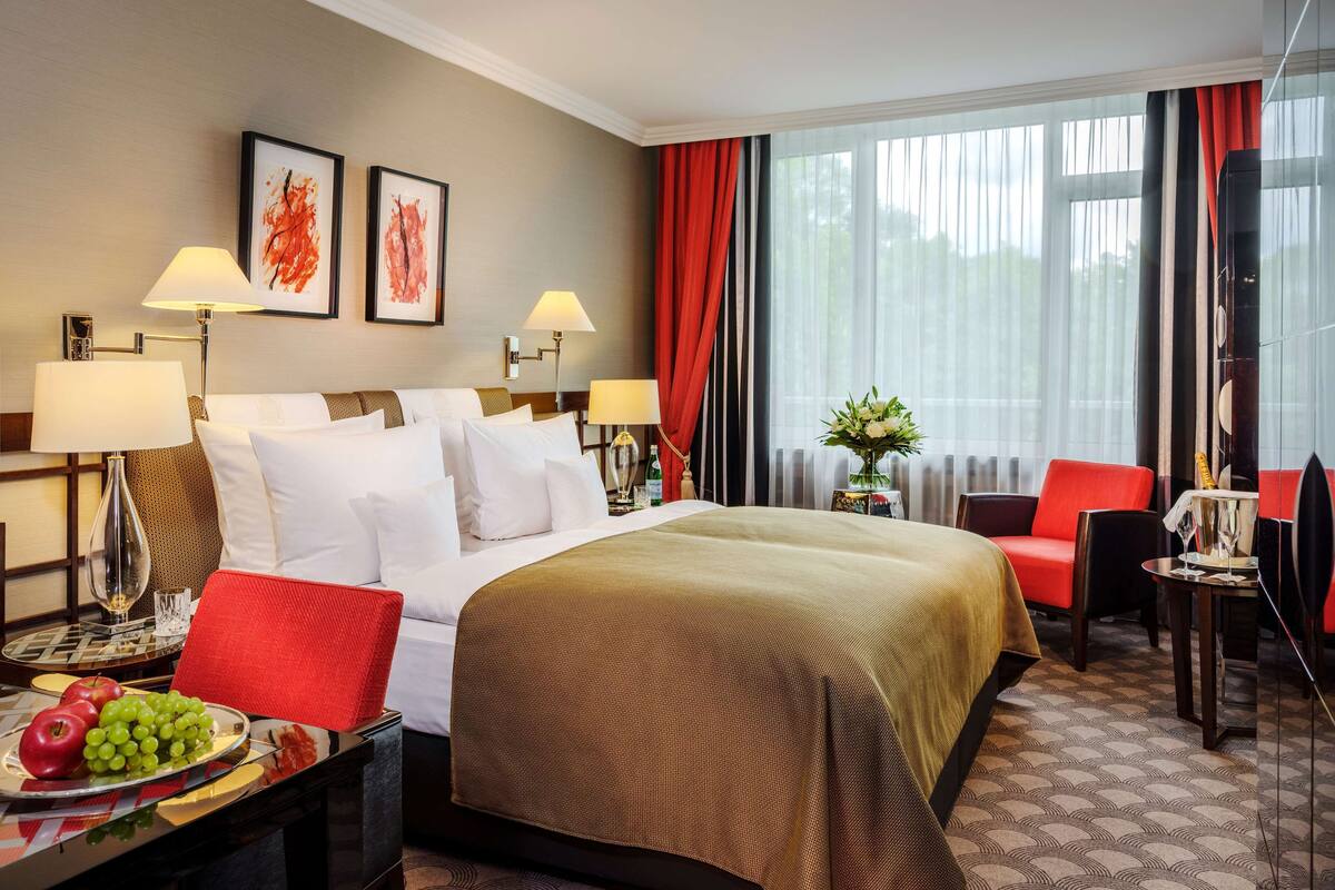 Quarto de hotel com uma cama grande, duas mesas de cabeceira com abajures, uma poltrona vermelha, uma pequena mesa com um vaso e obras de arte na parede.