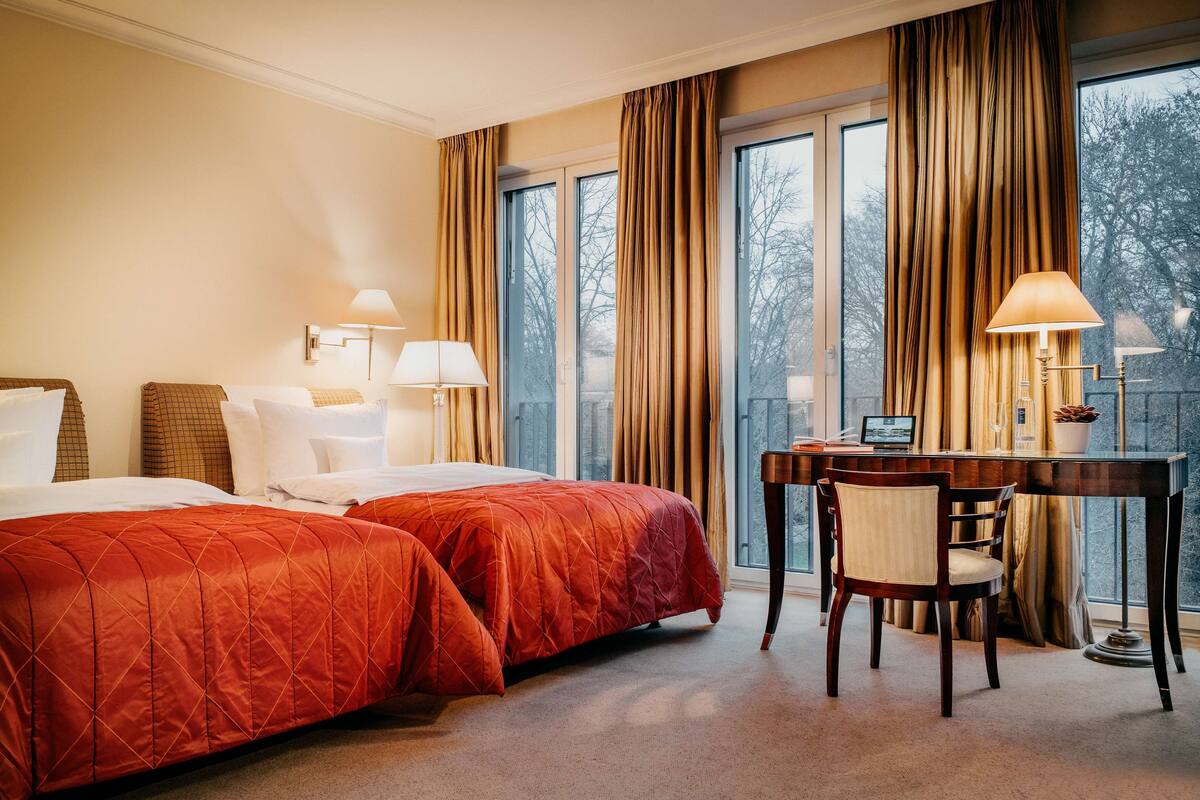 Quarto de hotel com duas camas, uma escrivaninha e uma cadeira. Há duas luminárias na escrivaninha e uma janela com cortinas.